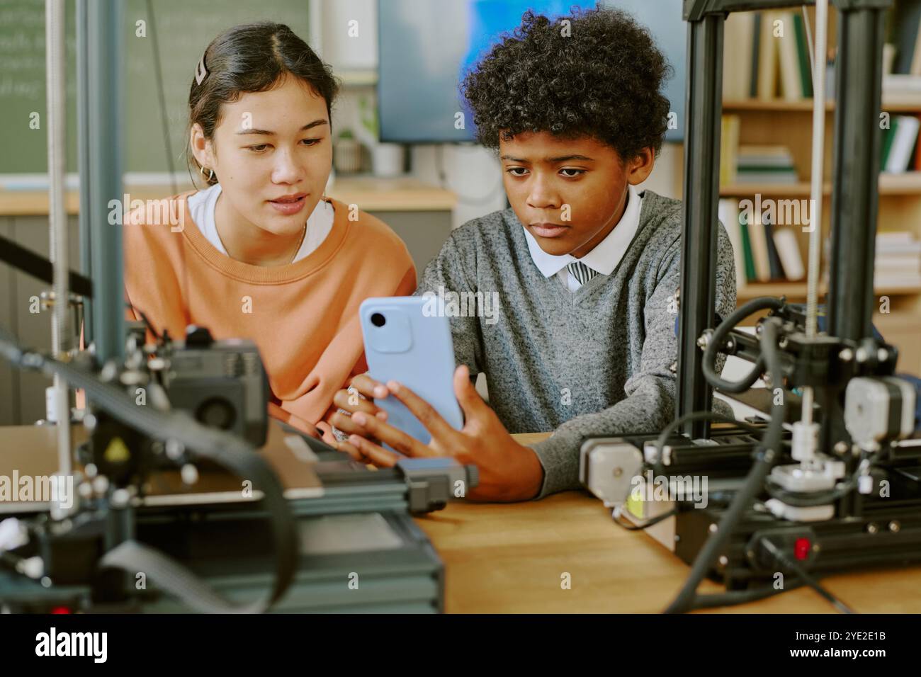Two students observing a 3D printer's progress in a classroom setting ...