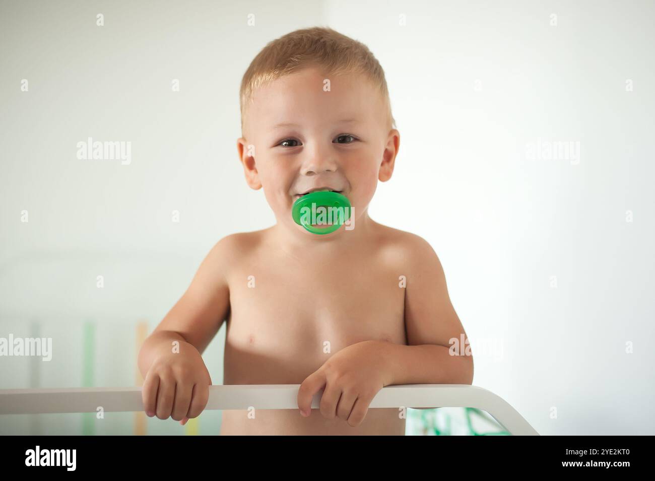 A young boy stands beside a crib, smiling and holding a green pacifier ...