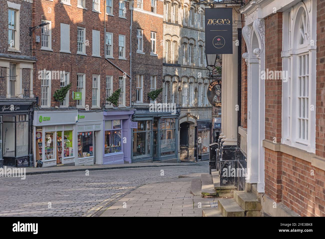 A section of an old cobbled street. Shops fronts line the pavement and ...