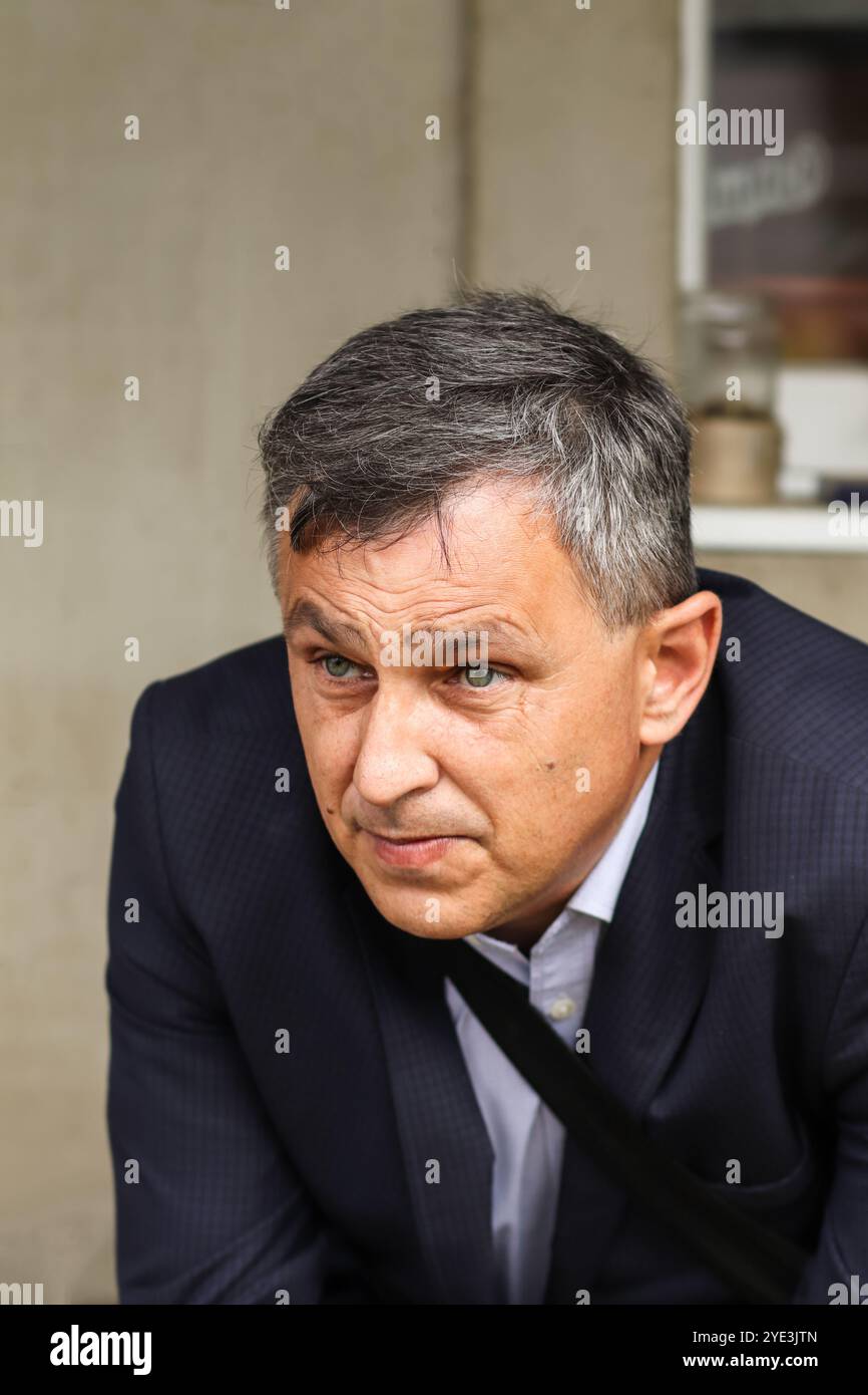 A serious man in his 40s or 50s, dressed in a tailored black suit ...