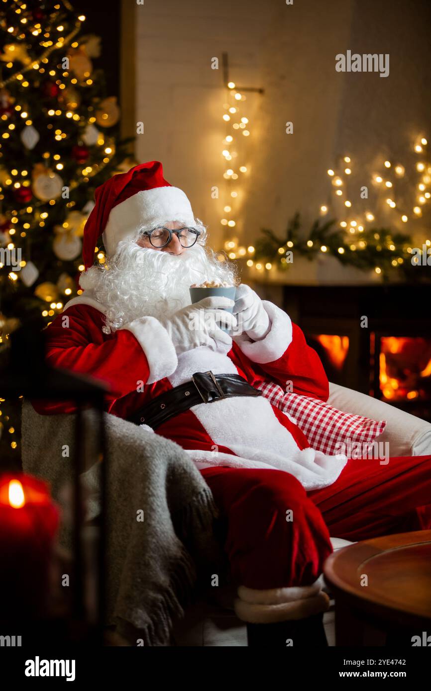 Jolly and relaxed Santa Claus with eyeglasses sits by fireplace, drinks ...