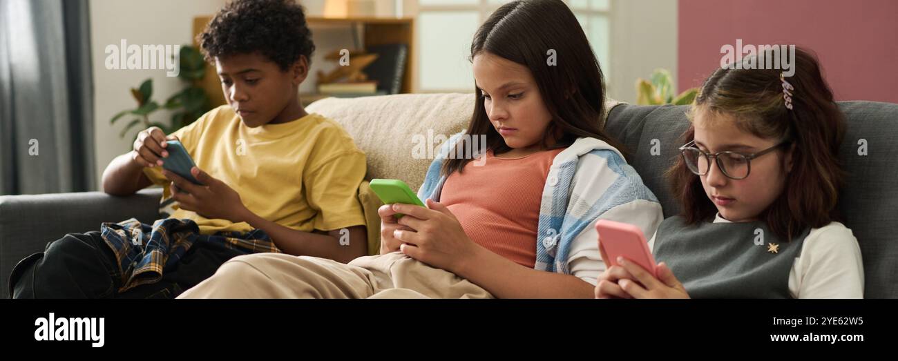 Group of three children sitting on sofa, each focused on their mobile ...