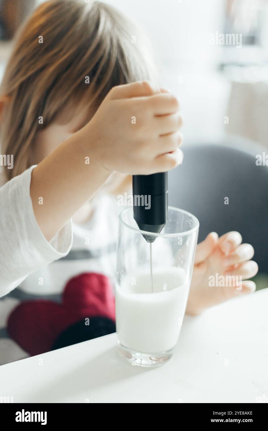 Pre-school child makes frothy milk with a hand mixer in a glass cup in ...