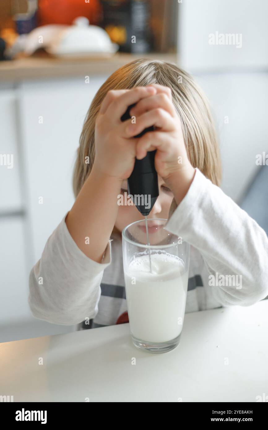 Pre-school child makes frothy milk with a hand mixer in a glass cup in ...