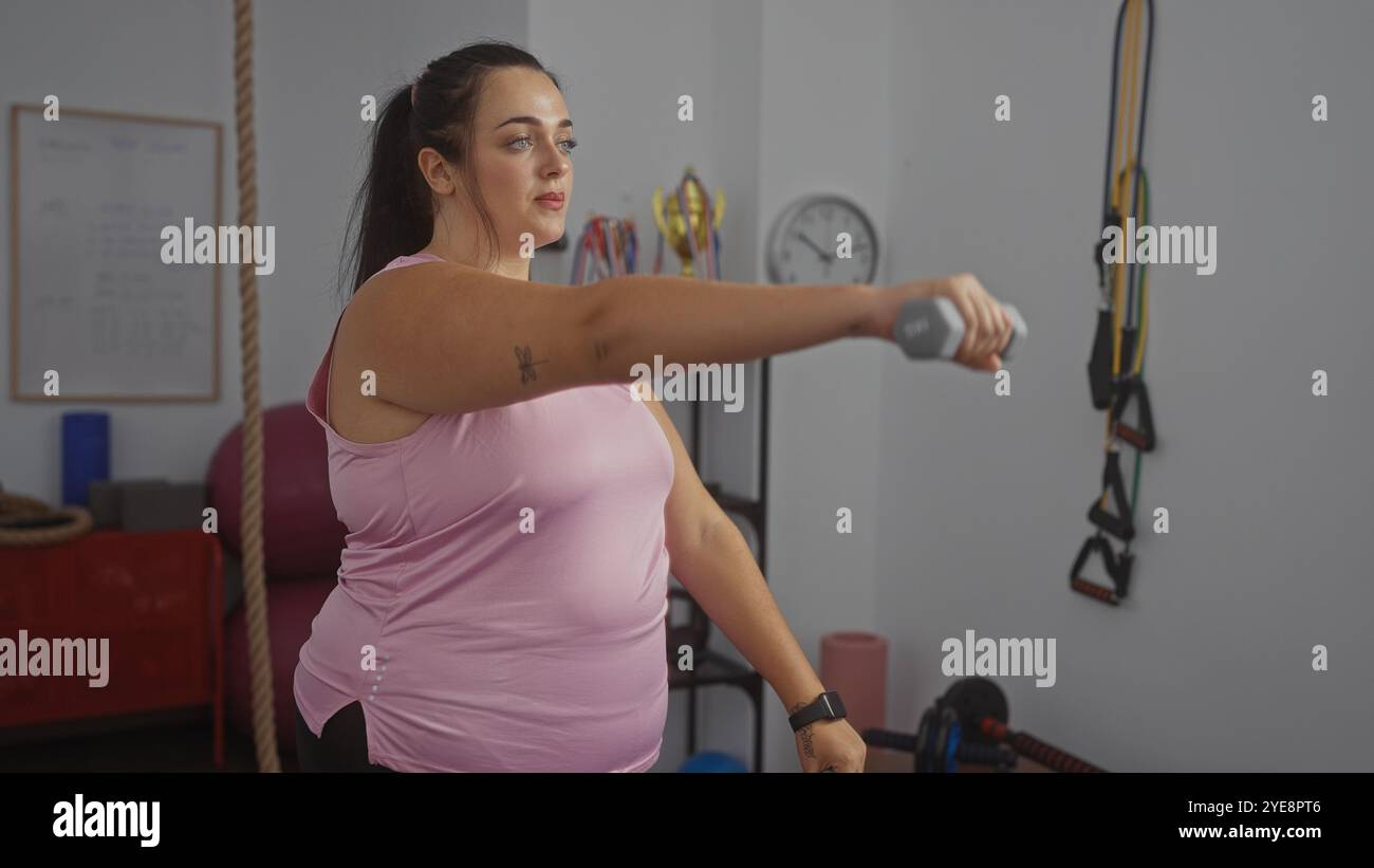Young curvy woman exercising indoors with a dumbbell in a gym setting, featuring fitness ...