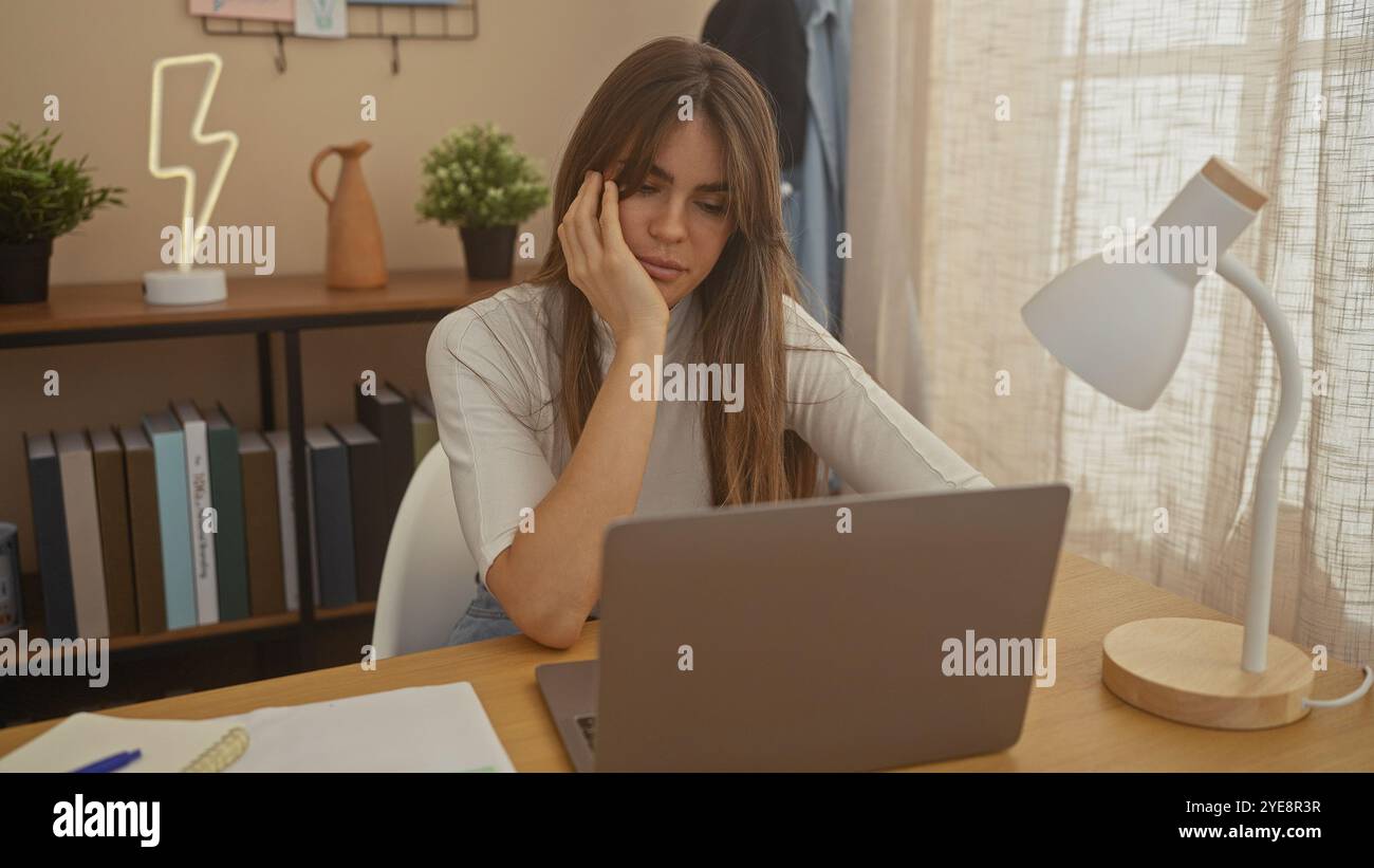 A thoughtful young woman working on a laptop in a well-lit home office ...