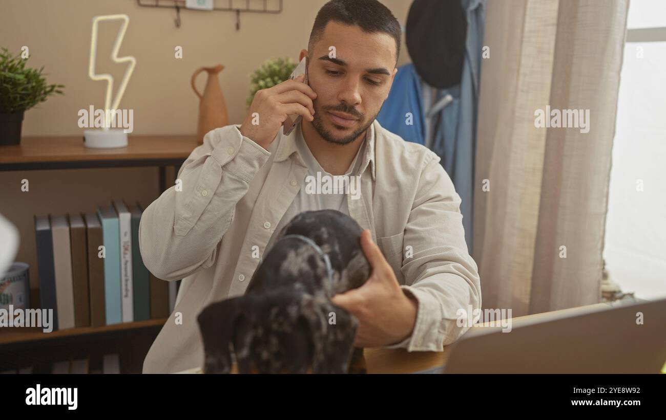 A handsome young hispanic man talks on the phone at home, while petting his dachshund in a cozy ...