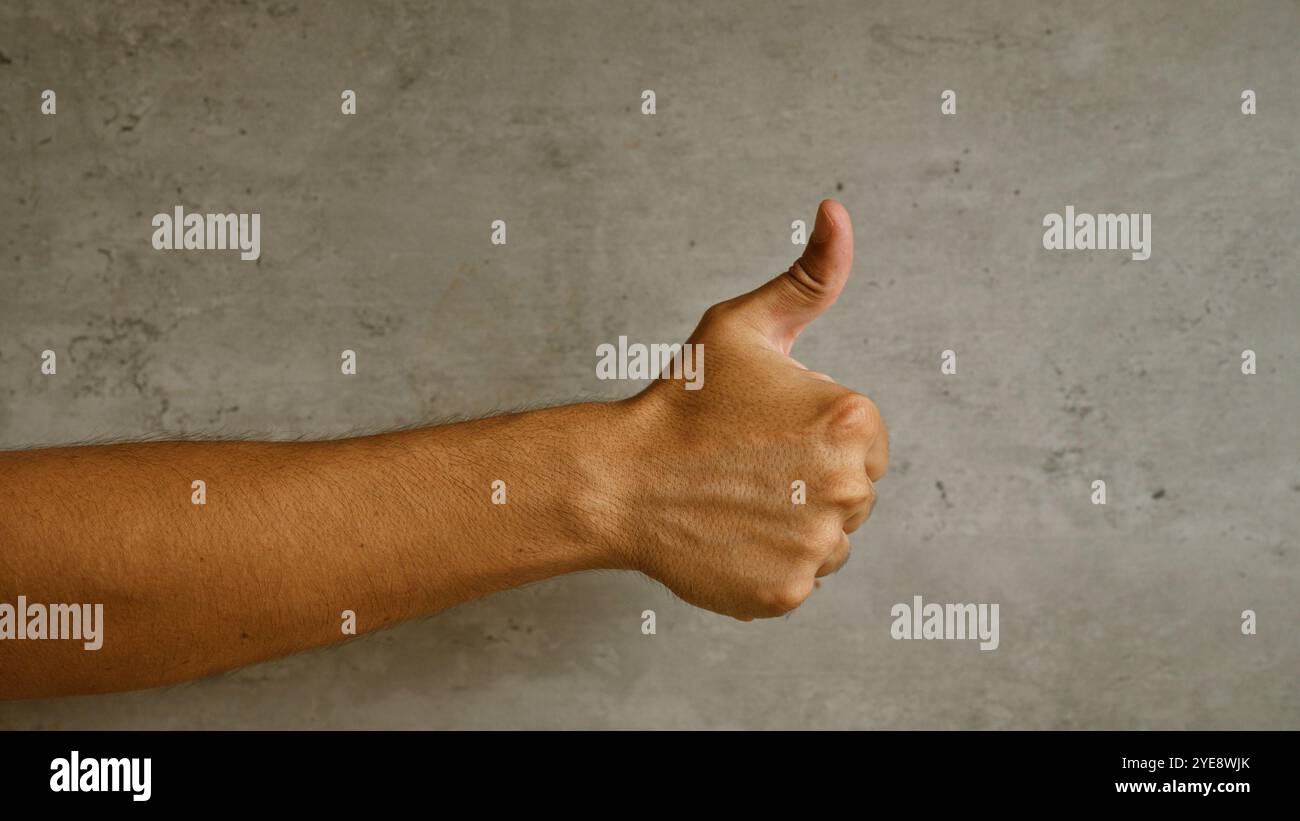 Man's hand giving a thumbs-up gesture against a concrete wall backdrop ...