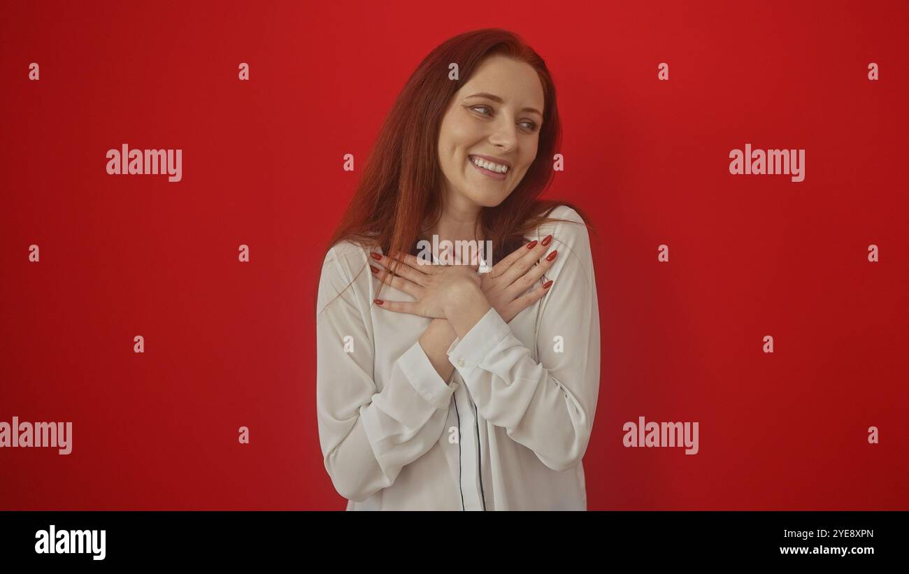A joyful redhead woman in a white blouse poses with a grateful gesture ...
