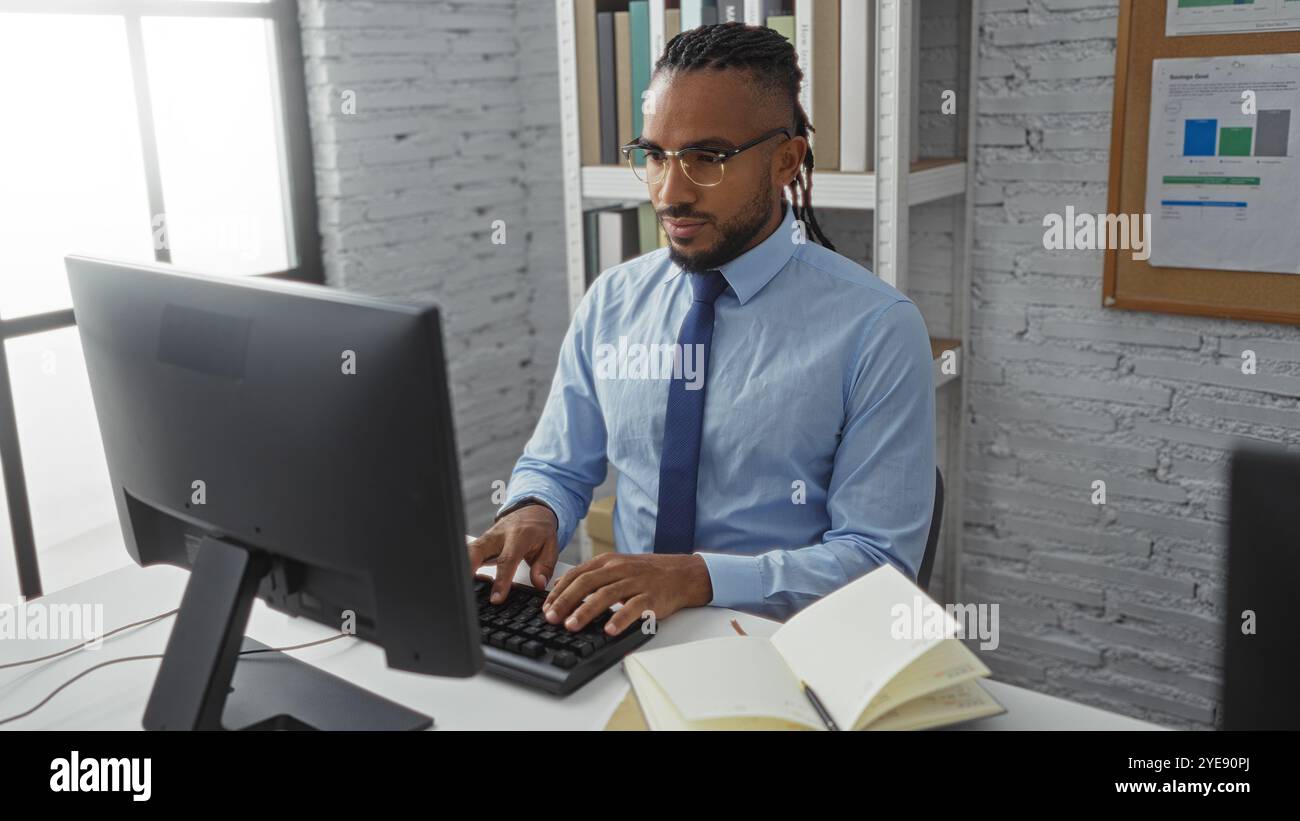Young man with braids working at desk in modern office with computer monitor, wearing glasses ...