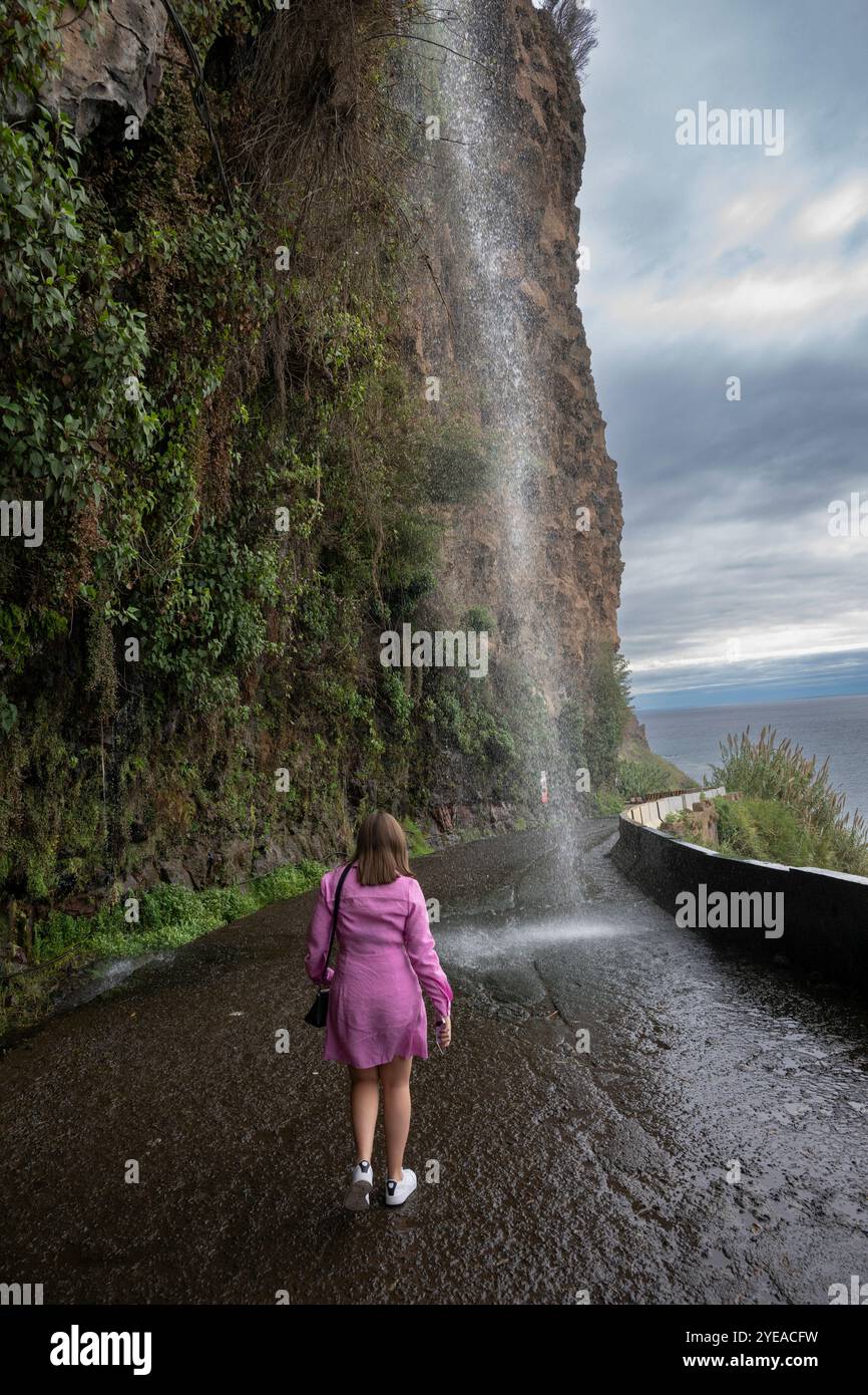 Woman stands watching a waterfall splashing down on a coastal roadway ...