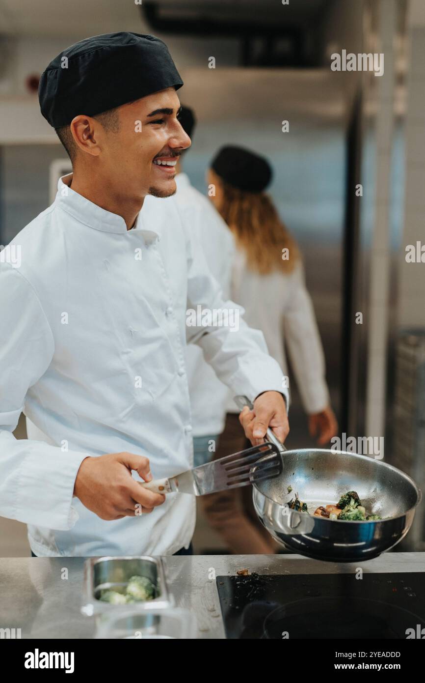 Happy male teenage chef trainee preparing food in saucepan at cooking ...