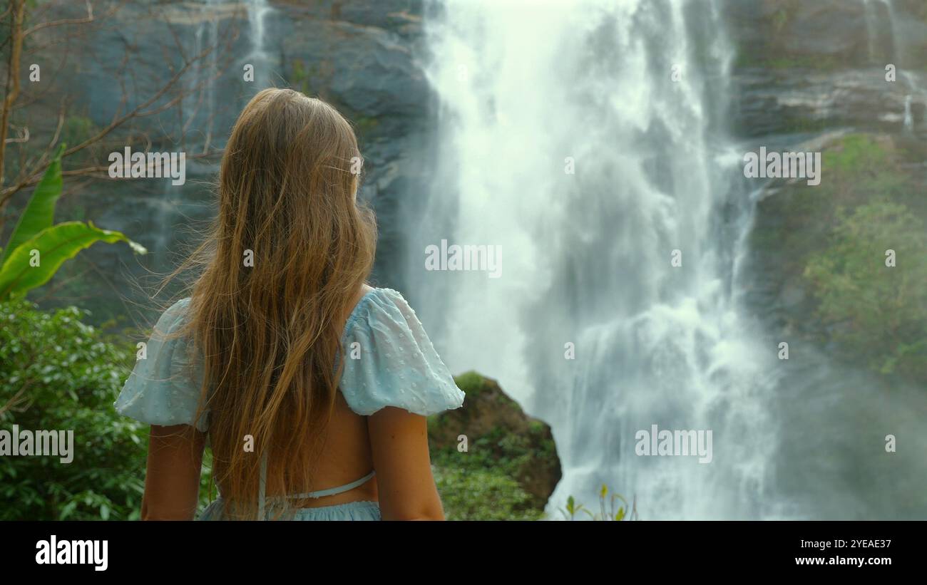 Young woman with long hair in dress stands facing majestic waterfall ...