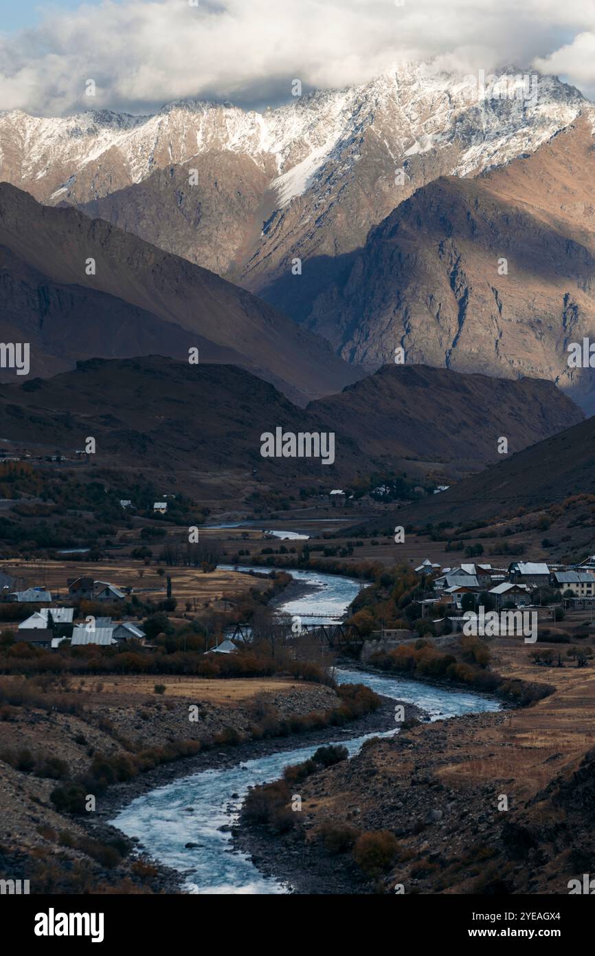 Village along a river in the remote valley below rugged snowy mountains ...