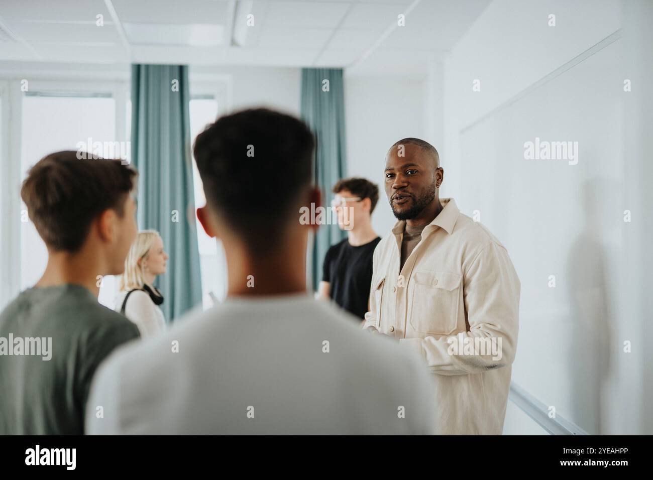 Male teacher discussing with students while standing by whiteboard in ...