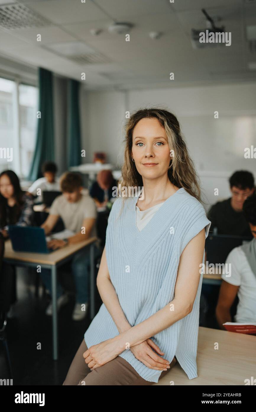 Portrait of female teacher sitting on desk in classroom at high school ...