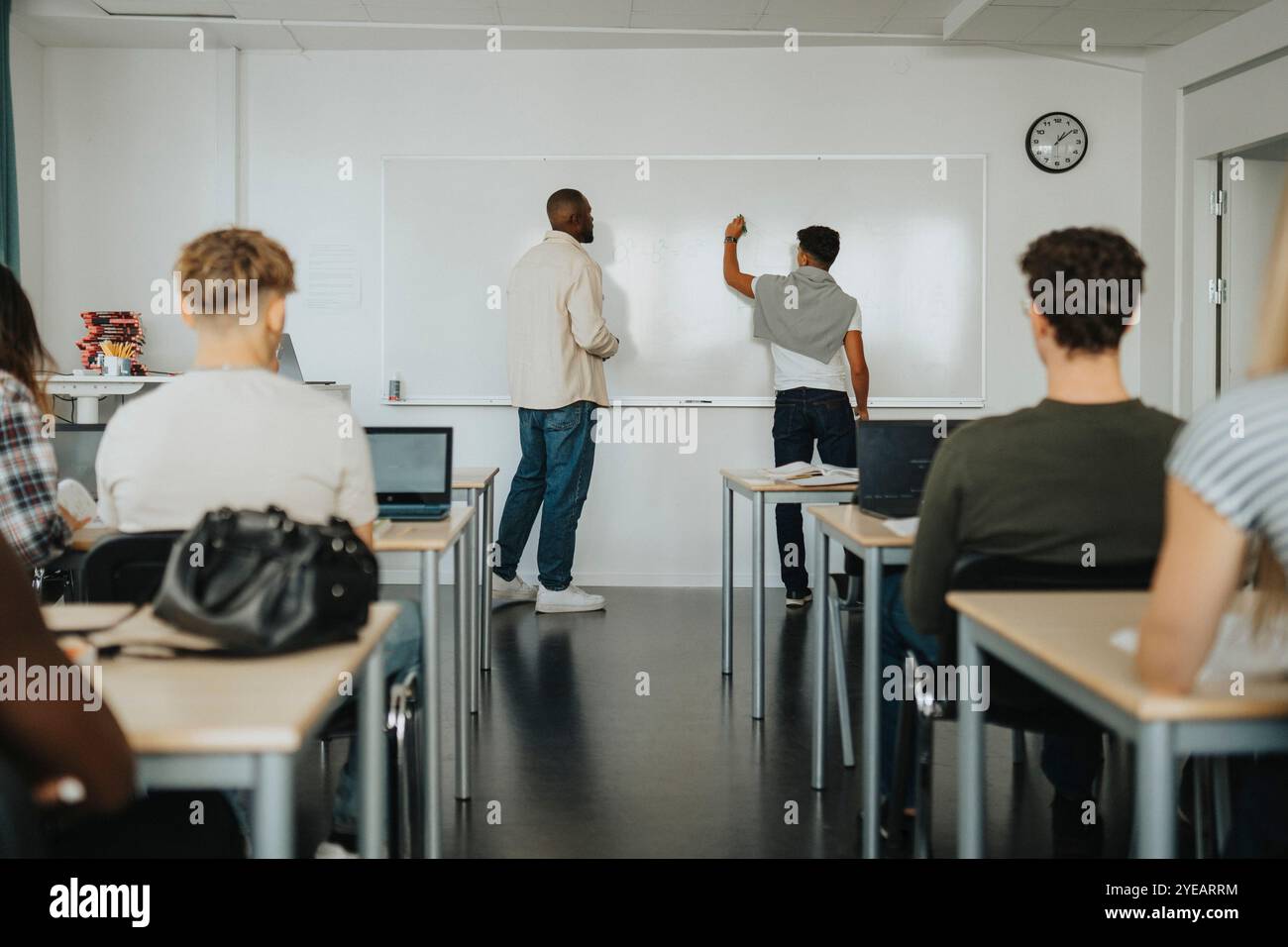 Rear view of male student writing on whiteboard by teacher standing ...