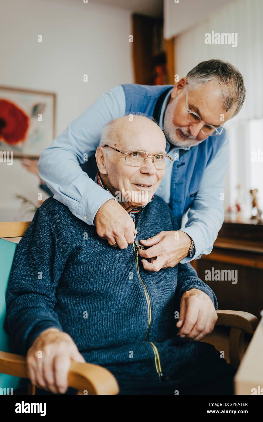 Senior man helping elderly friend in wearing jacket while sitting on ...