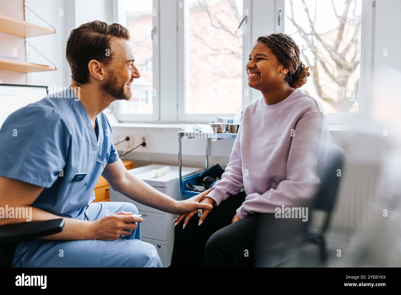 Happy male doctor consoling young women with hand on knee while sitting ...