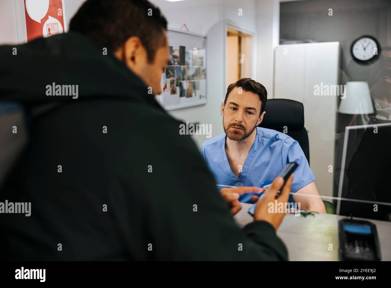 Male receptionist looking at patient's smart phone seen through glass ...