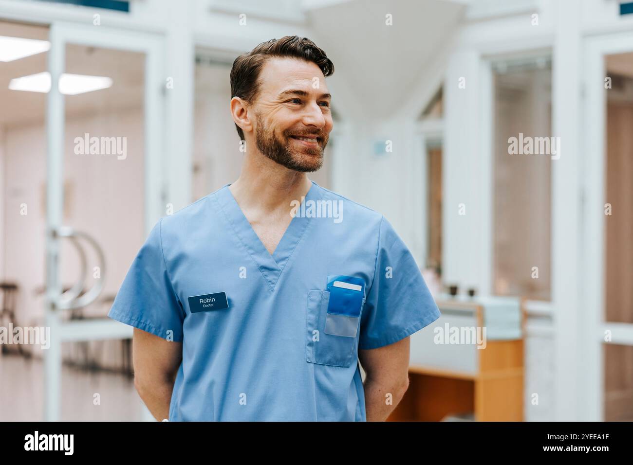 Happy male doctor wearing scrubs looking away with hands behind back in ...