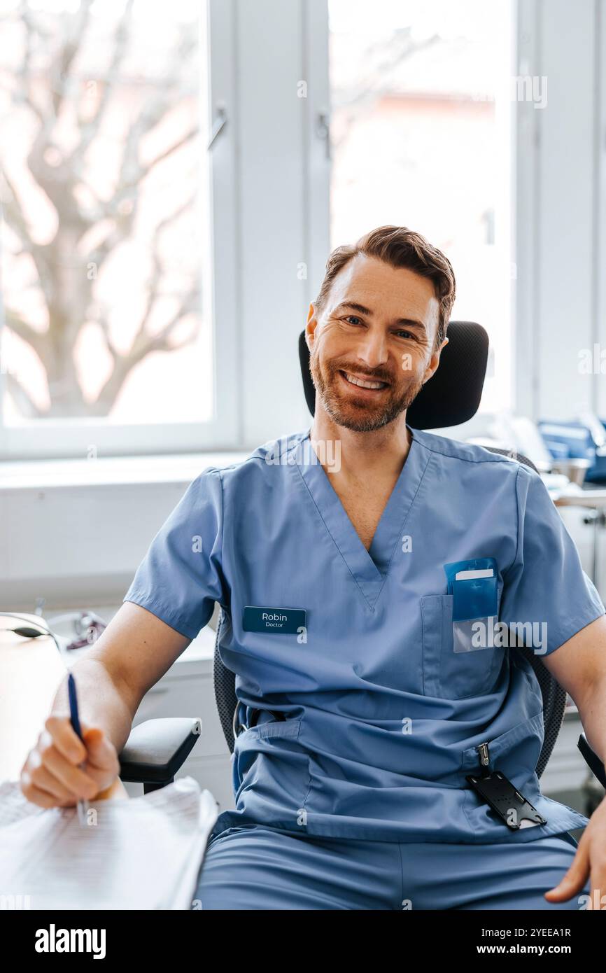 Portrait of happy male medical expert wearing scrubs holding pen and ...