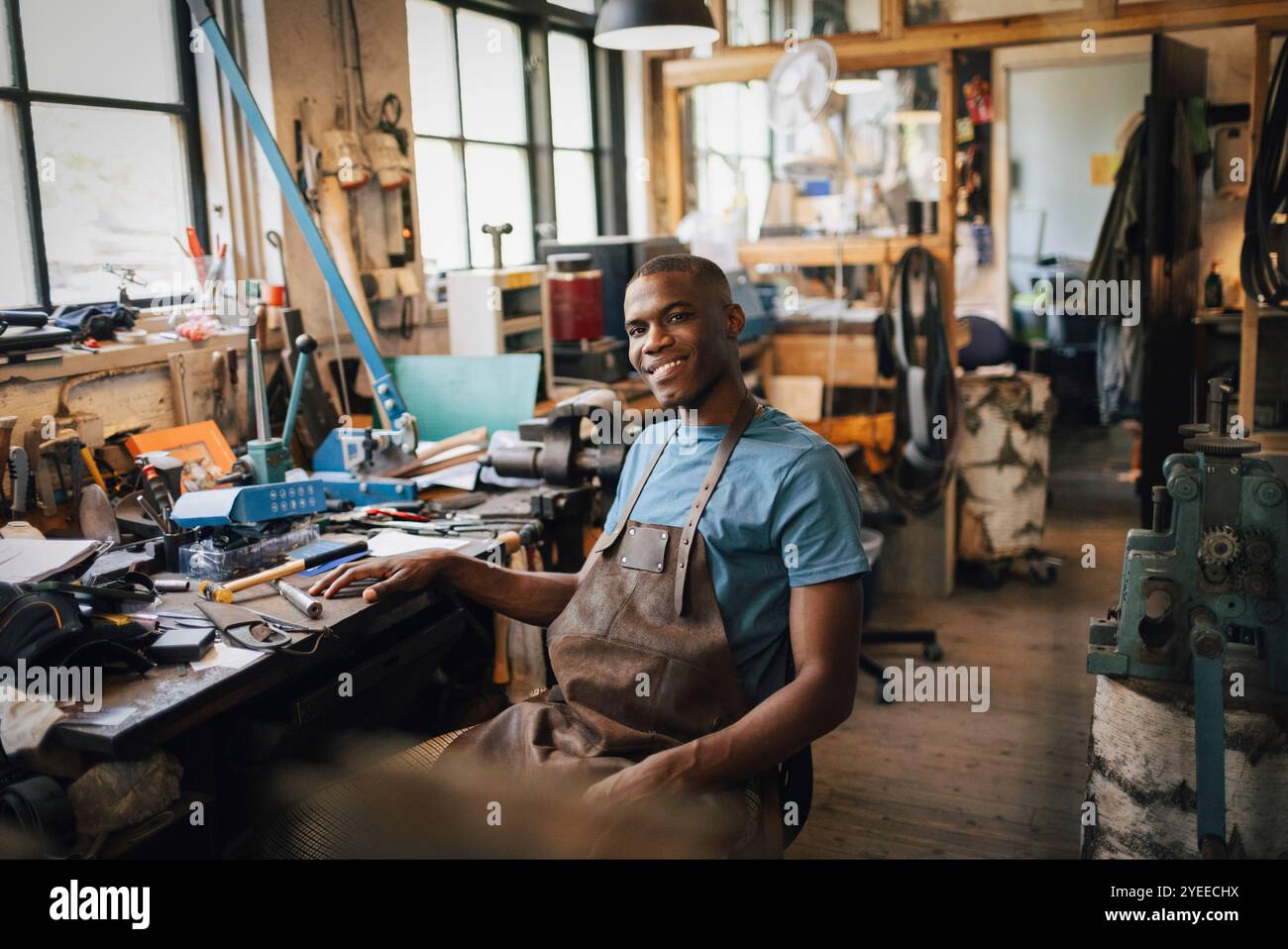 Smiling male craftsperson wearing apron and sitting on chair at ...