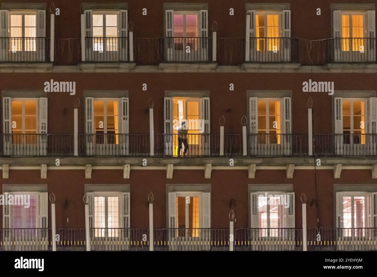 A nighttime view of an apartment building with lit windows and a person standing on a balcony ...