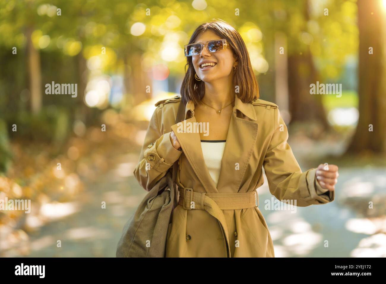 A woman wearing a tan trench coat and sunglasses is walking down a path ...