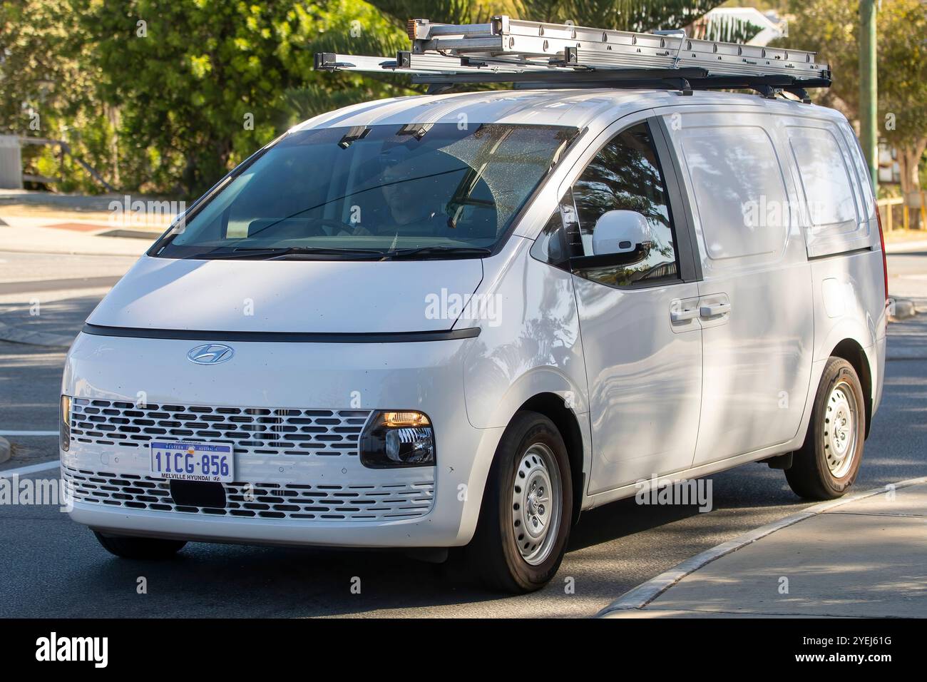 A white Hyundai Staria Cargo US4 van driving on a suburban street with a rooftop ladder rack ...
