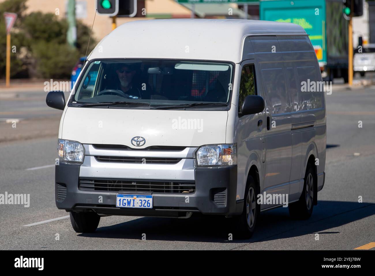 The photo shows a white Toyota HiAce van driving along a city street ...
