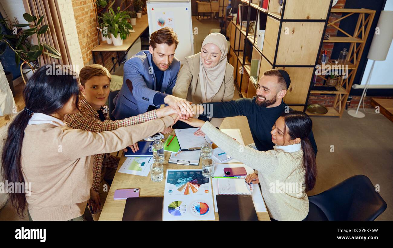 Group of multicultural colleagues join hands in team huddle around desk, symbolizing unity and ...