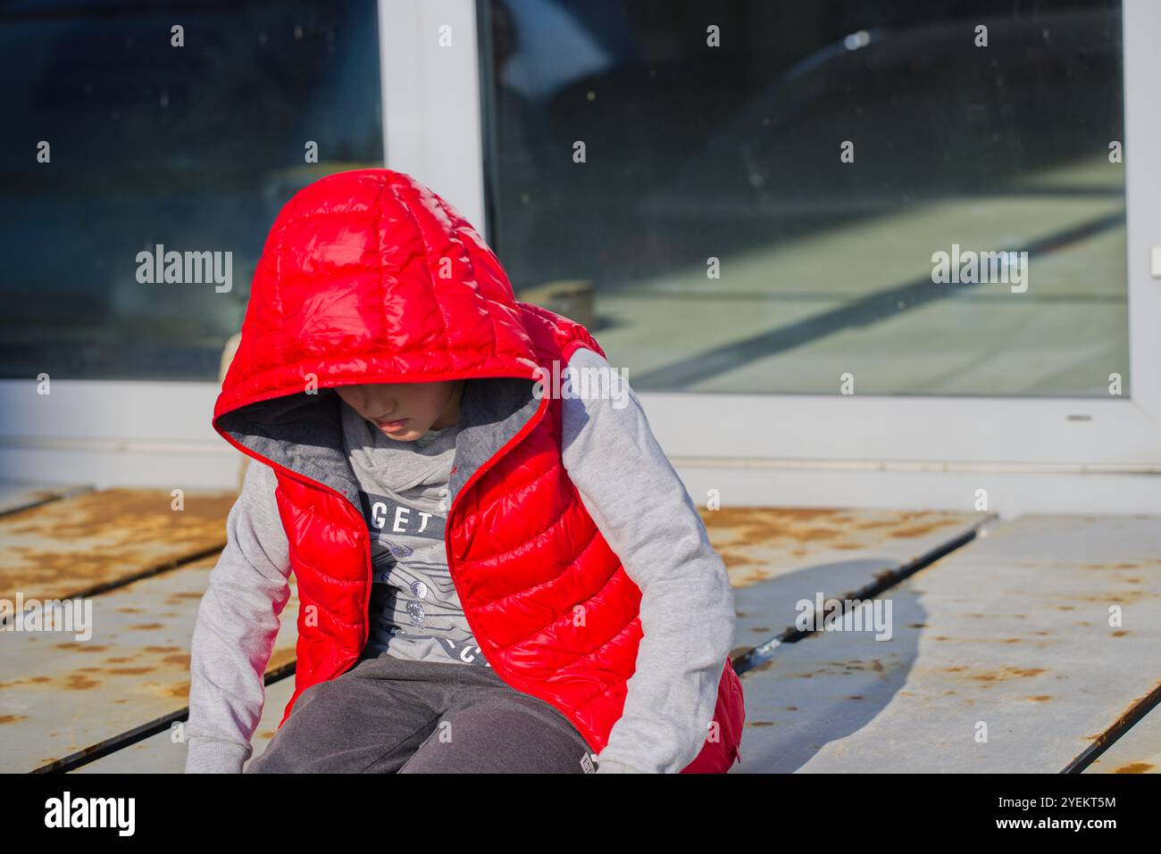 A boy in a red hooded vest sits alone on a sunny day, head lowered in ...