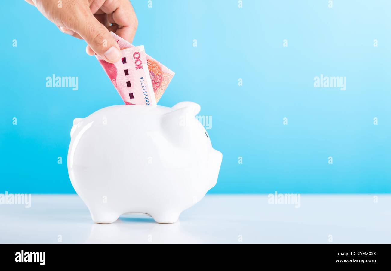 Hand holding a 100 Yuan note, dropping it into a white piggy bank, with ...