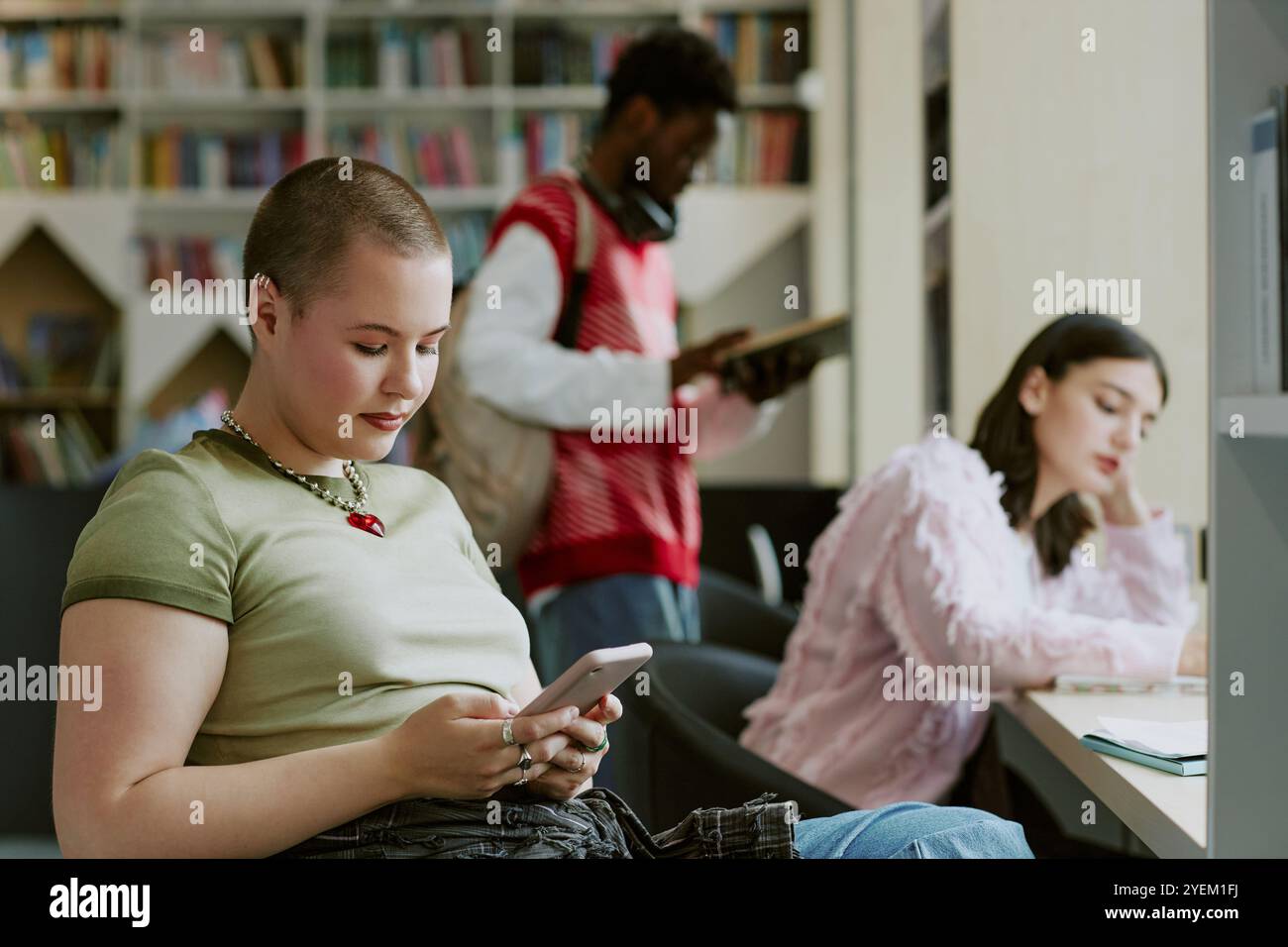 Students studying in a library setting with bookshelves in background, using smartphones and ...