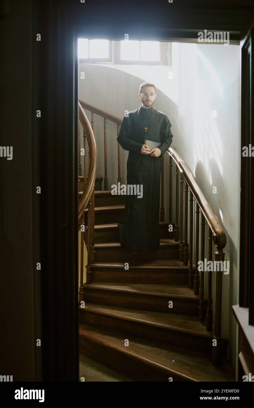 Vertical shot of young priest holding Bible in black cover standing on ...
