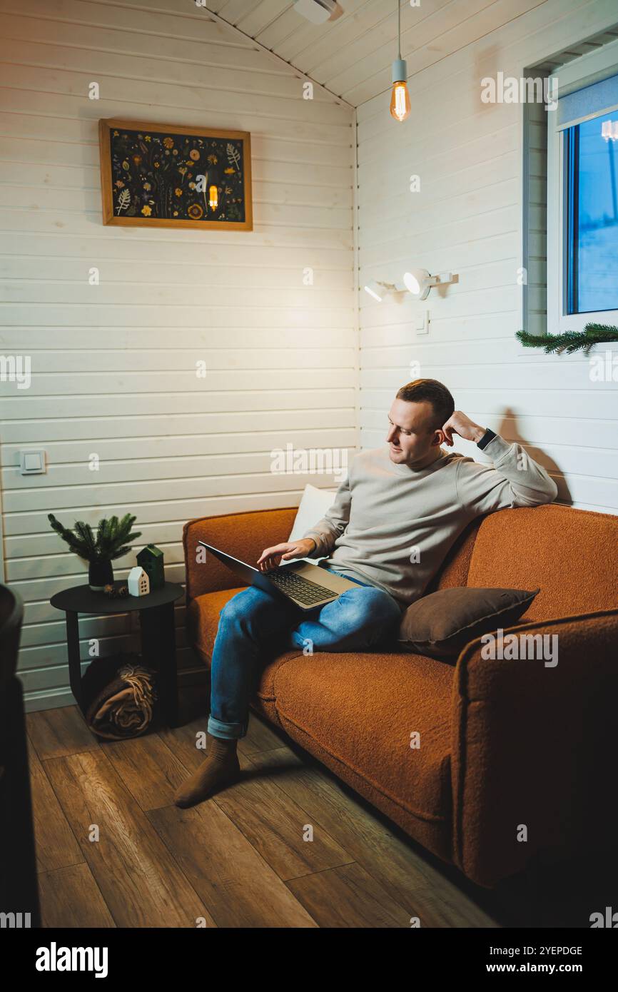 A handsome man in home clothes sits on a sofa and works on a laptop ...