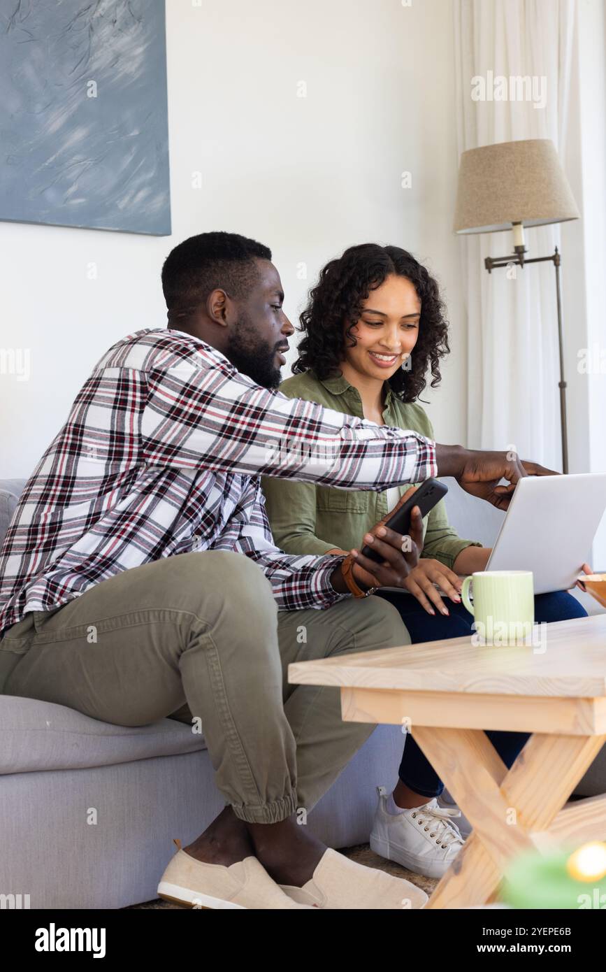 multiracial couple using laptop and smartphone at home, smiling and ...