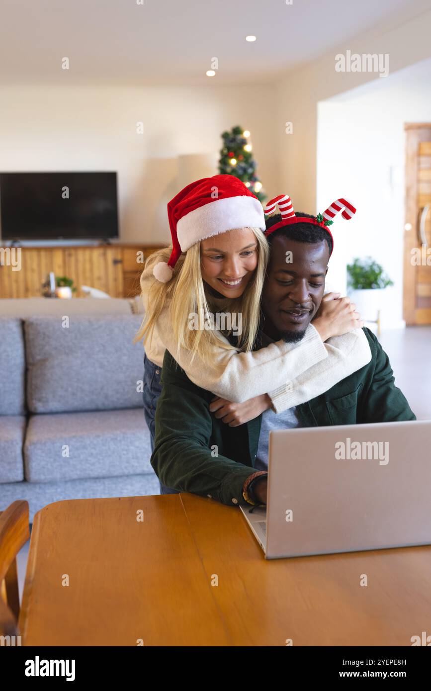 Happy multiracial couple wearing festive hats using laptop at home ...