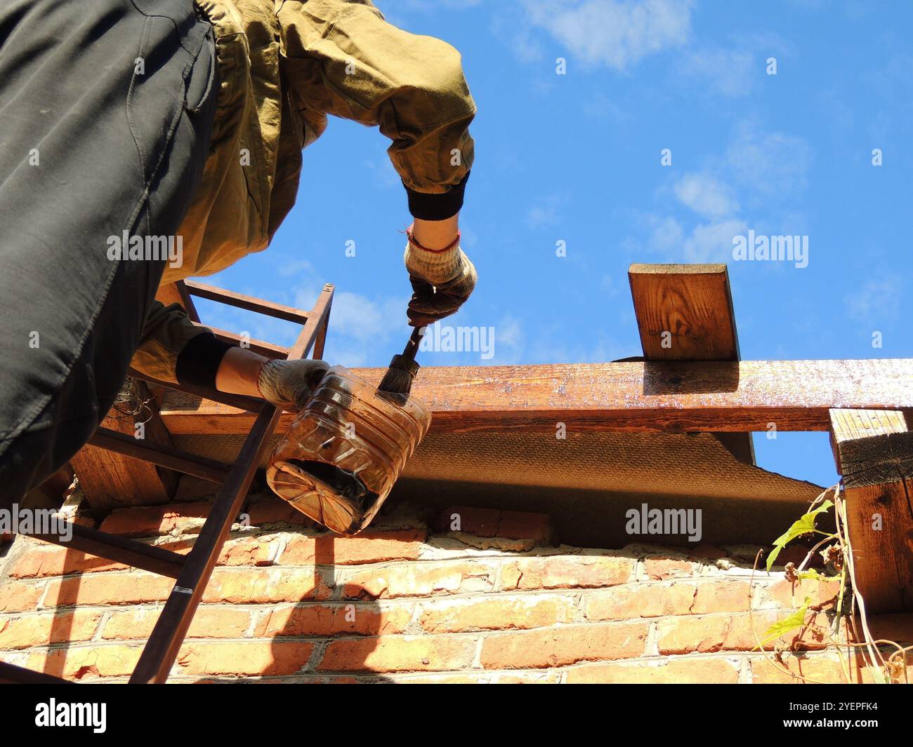 A man in work clothes and gloves paints or brushes a wooden roof ...
