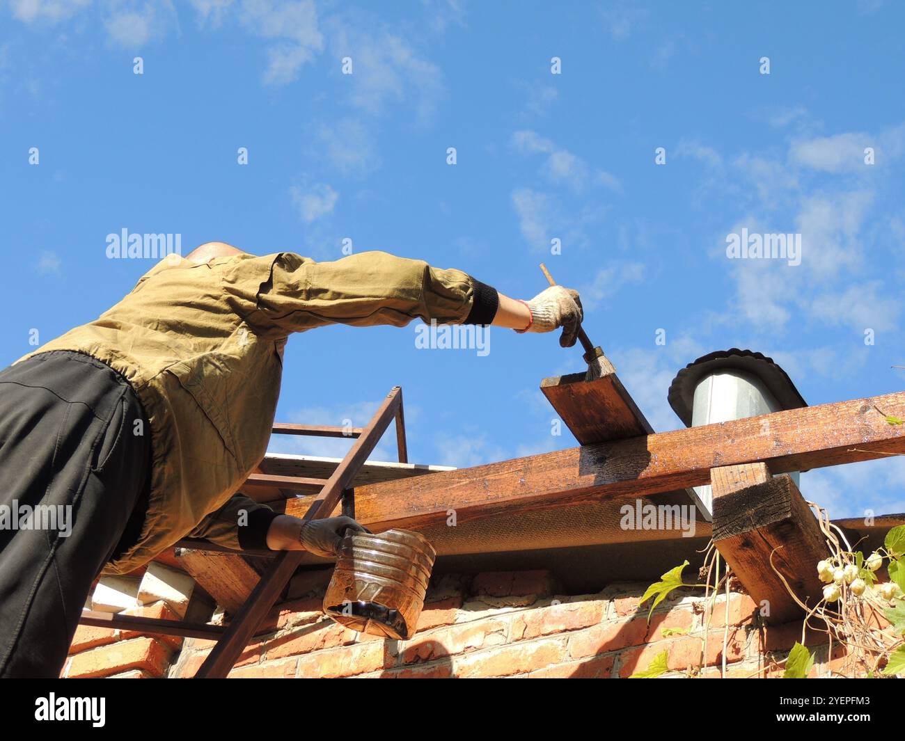 A man in work clothes and gloves paints or brushes a wooden roof ...