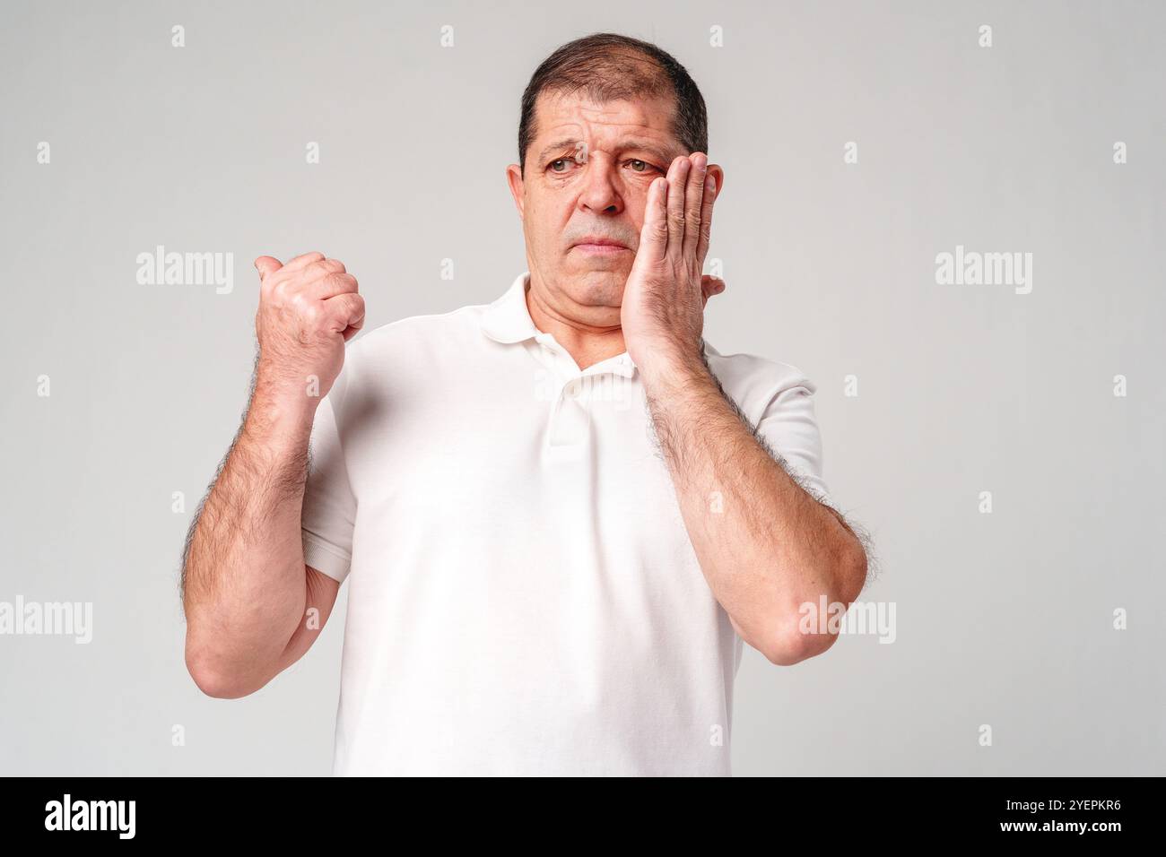 An older man holds his cheek, showing concern, against a plain ...