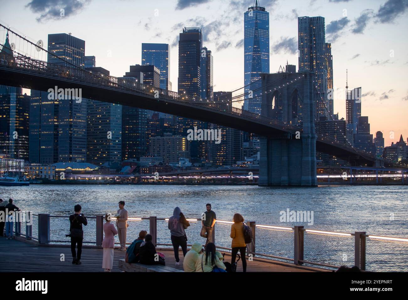Brooklyn Bridge with the Manhattan skyline at dusk. People gather on the waterfront, enjoying ...