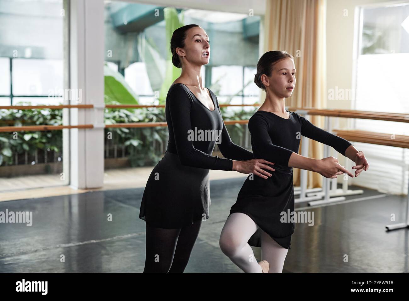 Graceful female dance teacher dressed in black leotard supporting little concentrated girl with ...