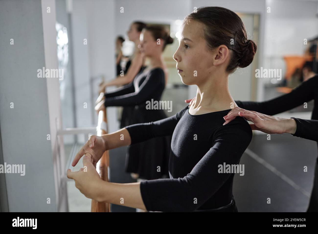 Side view of young girl standing at barre with teachers hands on ...