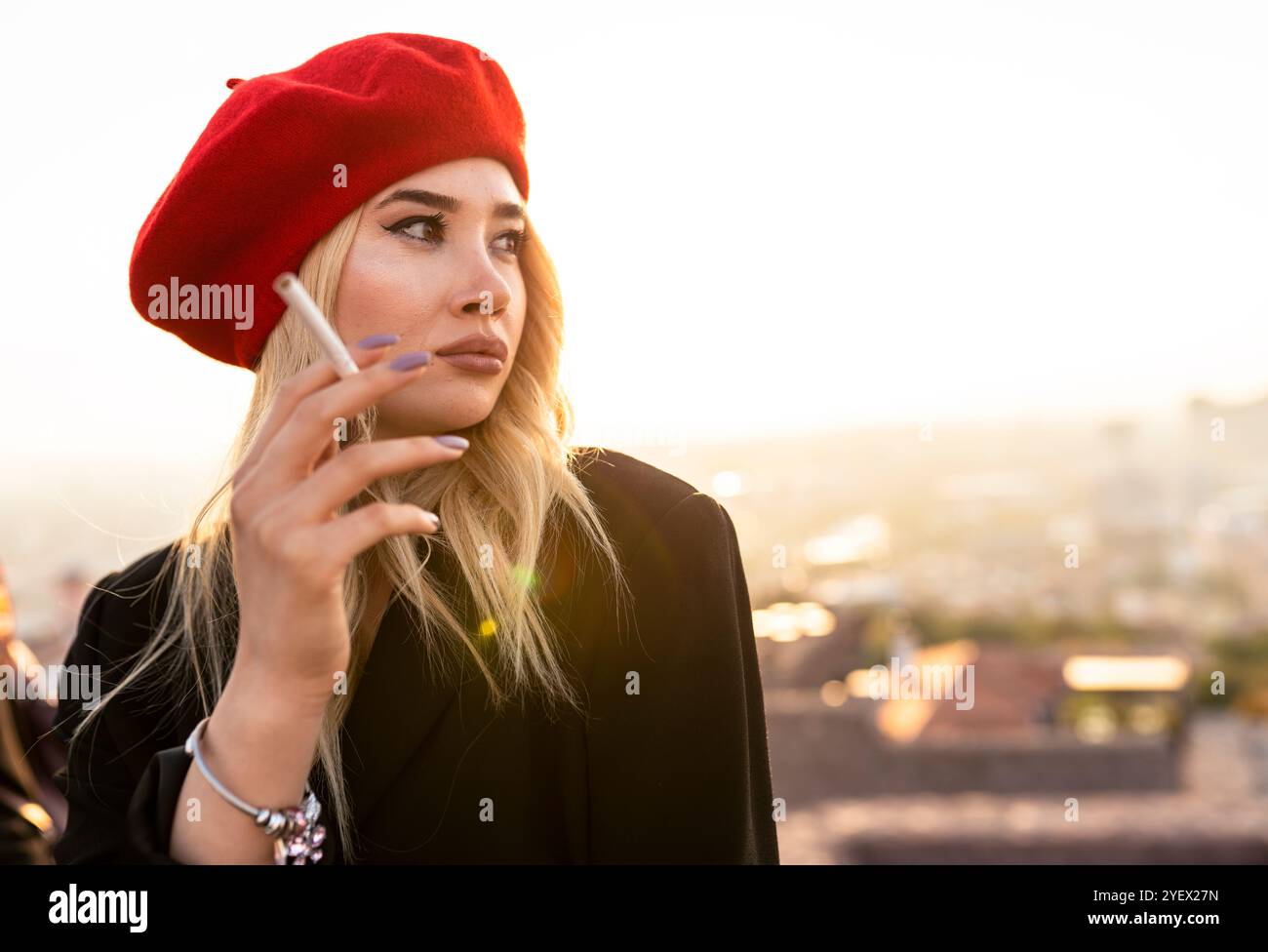 A woman in a red hat smokes a cigarette while looking out over a city ...