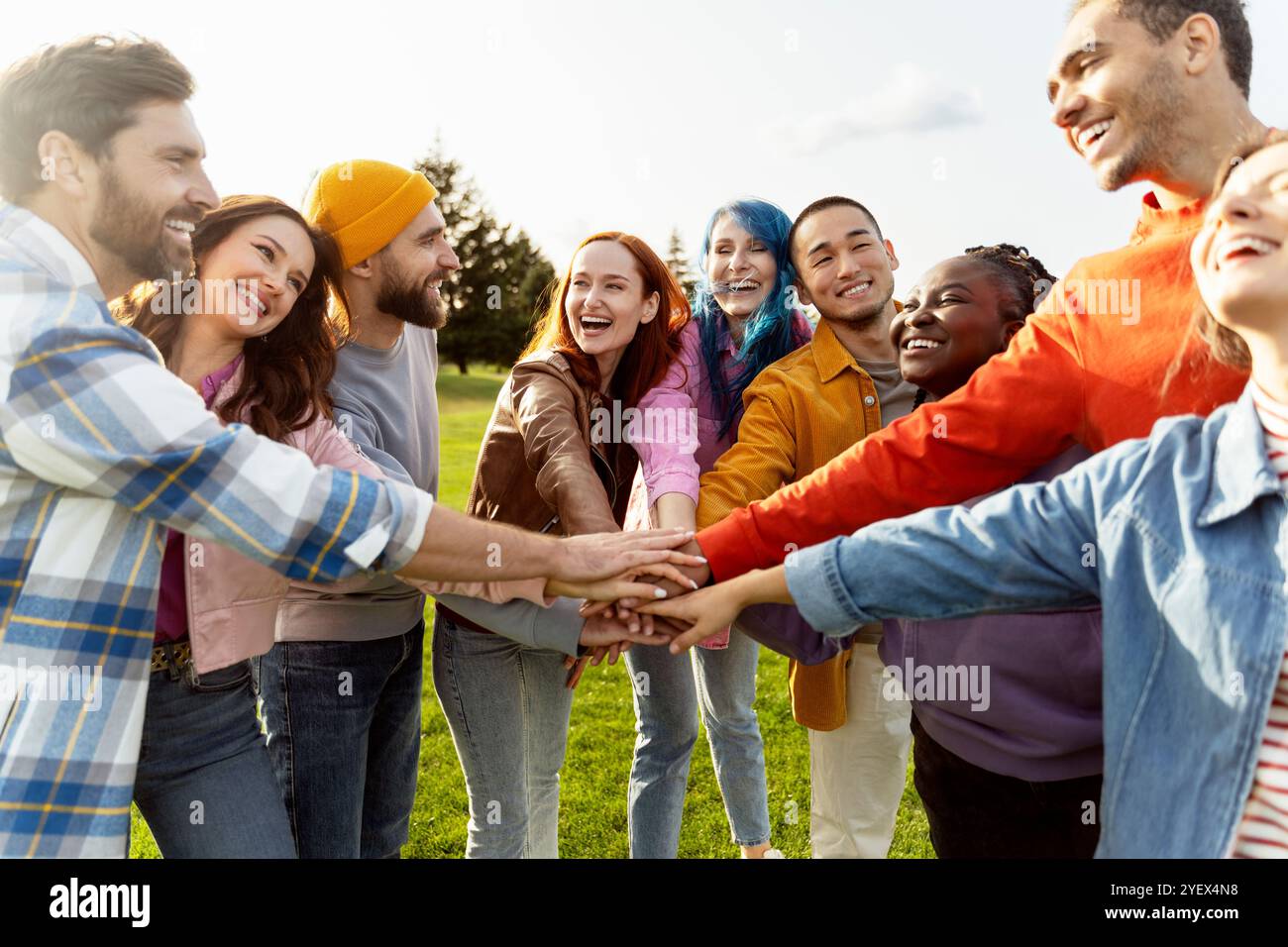 Group of diverse young people are standing in a circle on a green lawn, joining their hands ...
