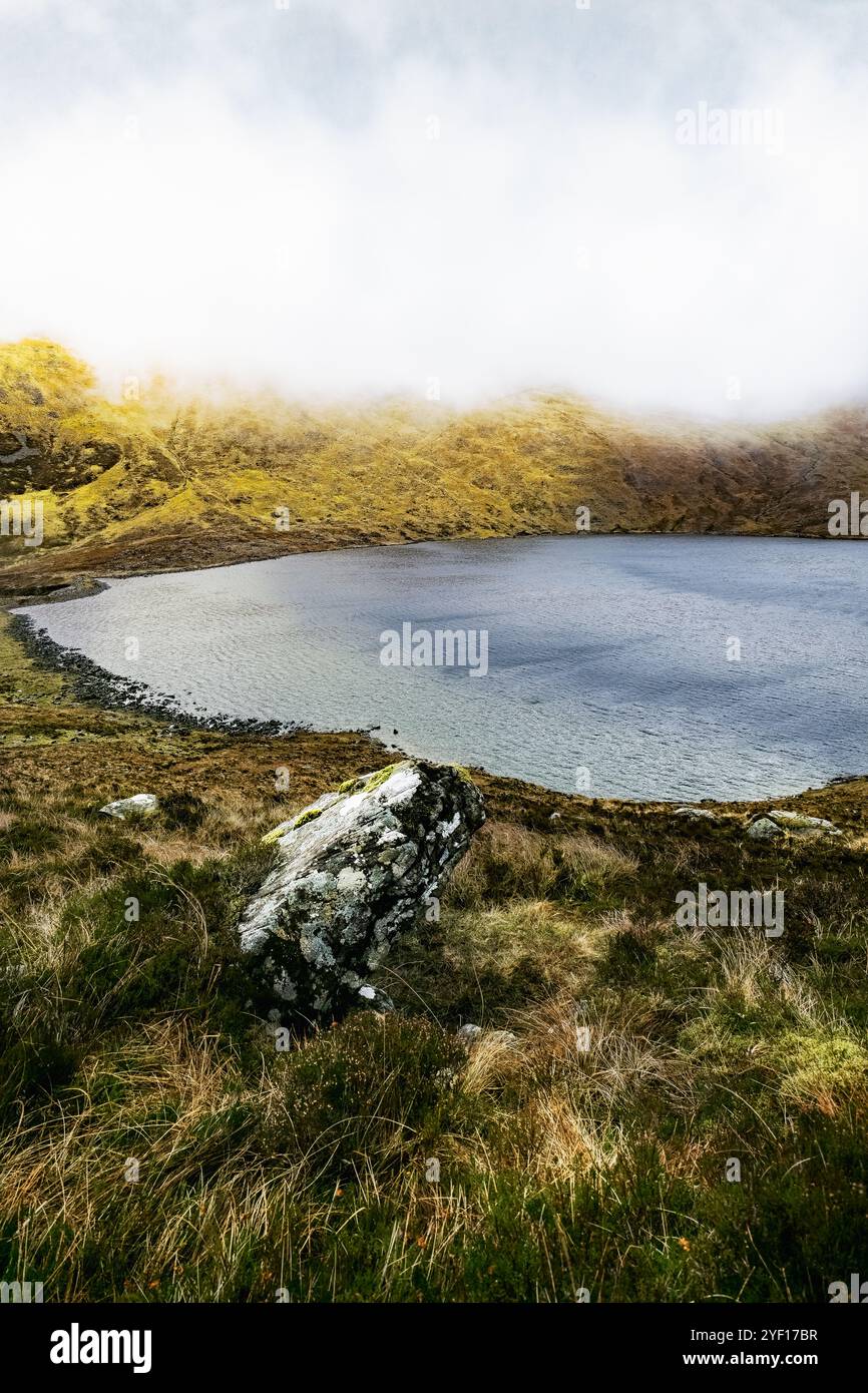 A peaceful mountain lake reflects the surrounding hills, partially ...