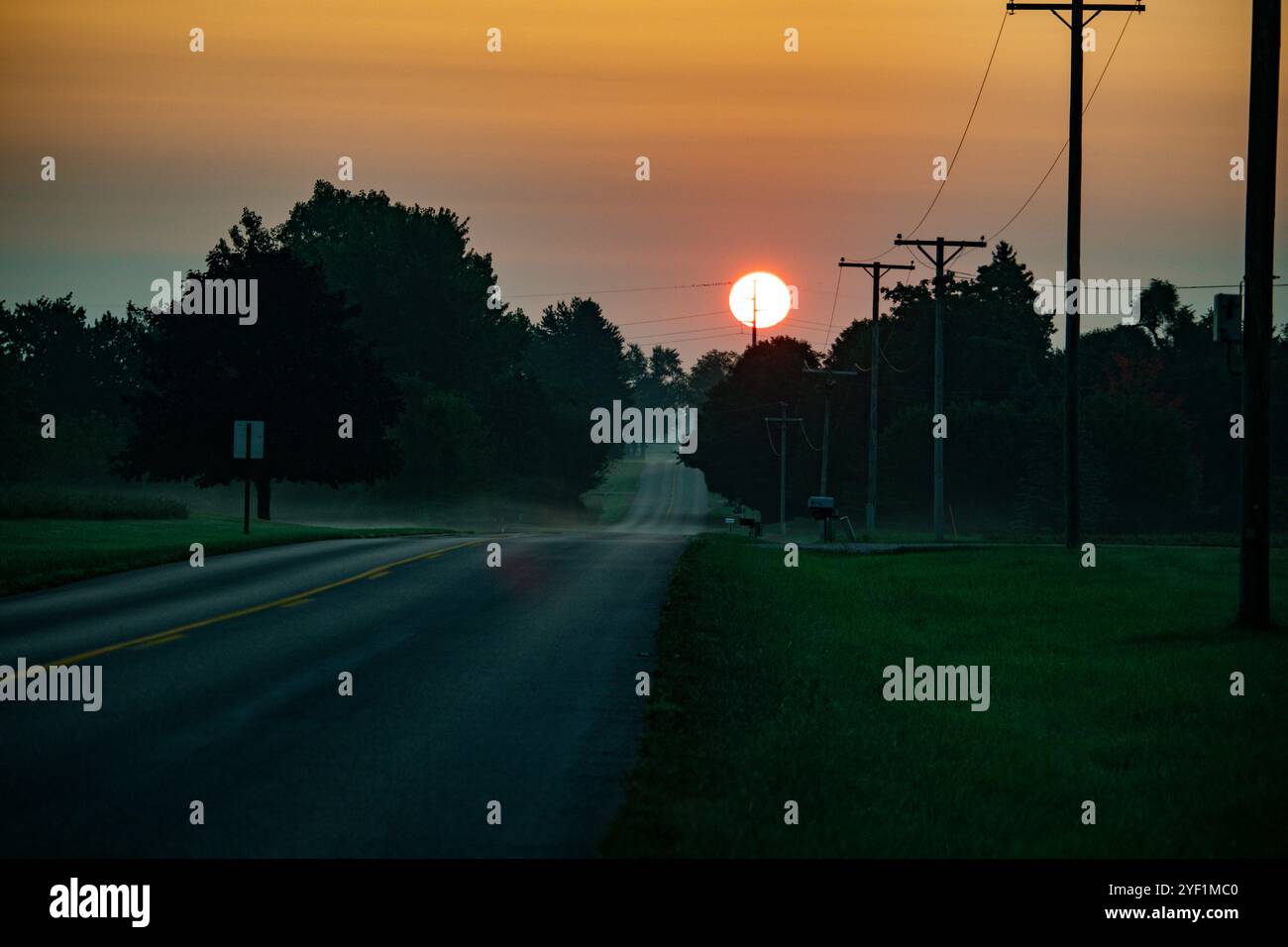 Early morning sunrise on a country road with ground fog and dewy grass ...