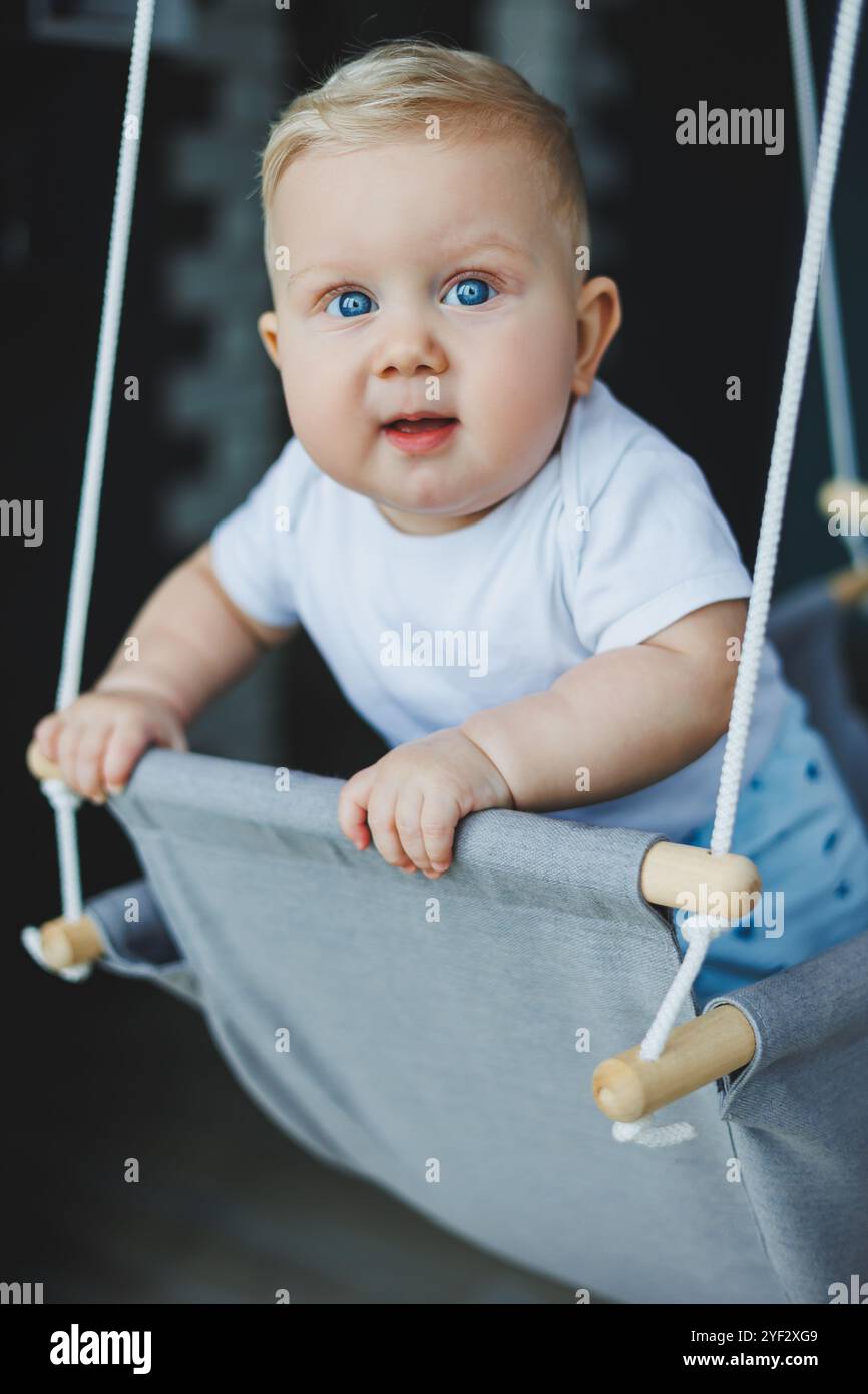 Baby boy swings in a swing at home. The child learns to have fun. Child ...